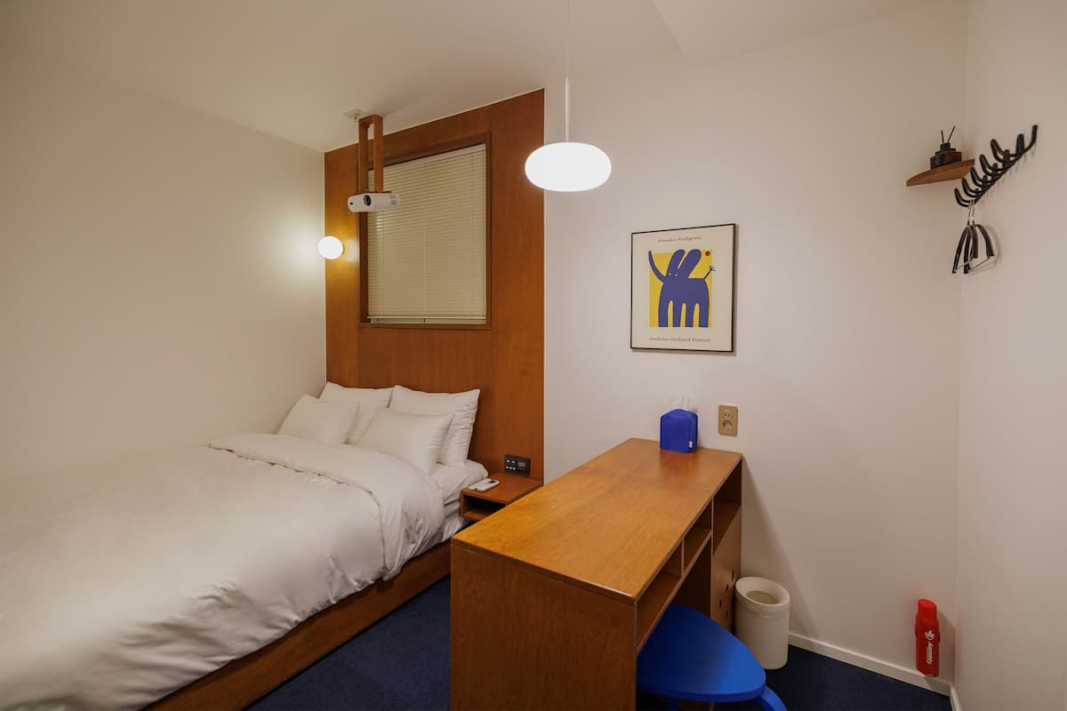 Quarto com cama, escrivaninha, cadeira, lixeira, quadro na parede e estante com objetos.