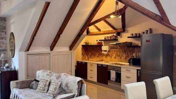 Quarto estilo loft com teto alto, vigas de madeira aparentes, sofá, mesa de jantar e cozinha com claraboia.