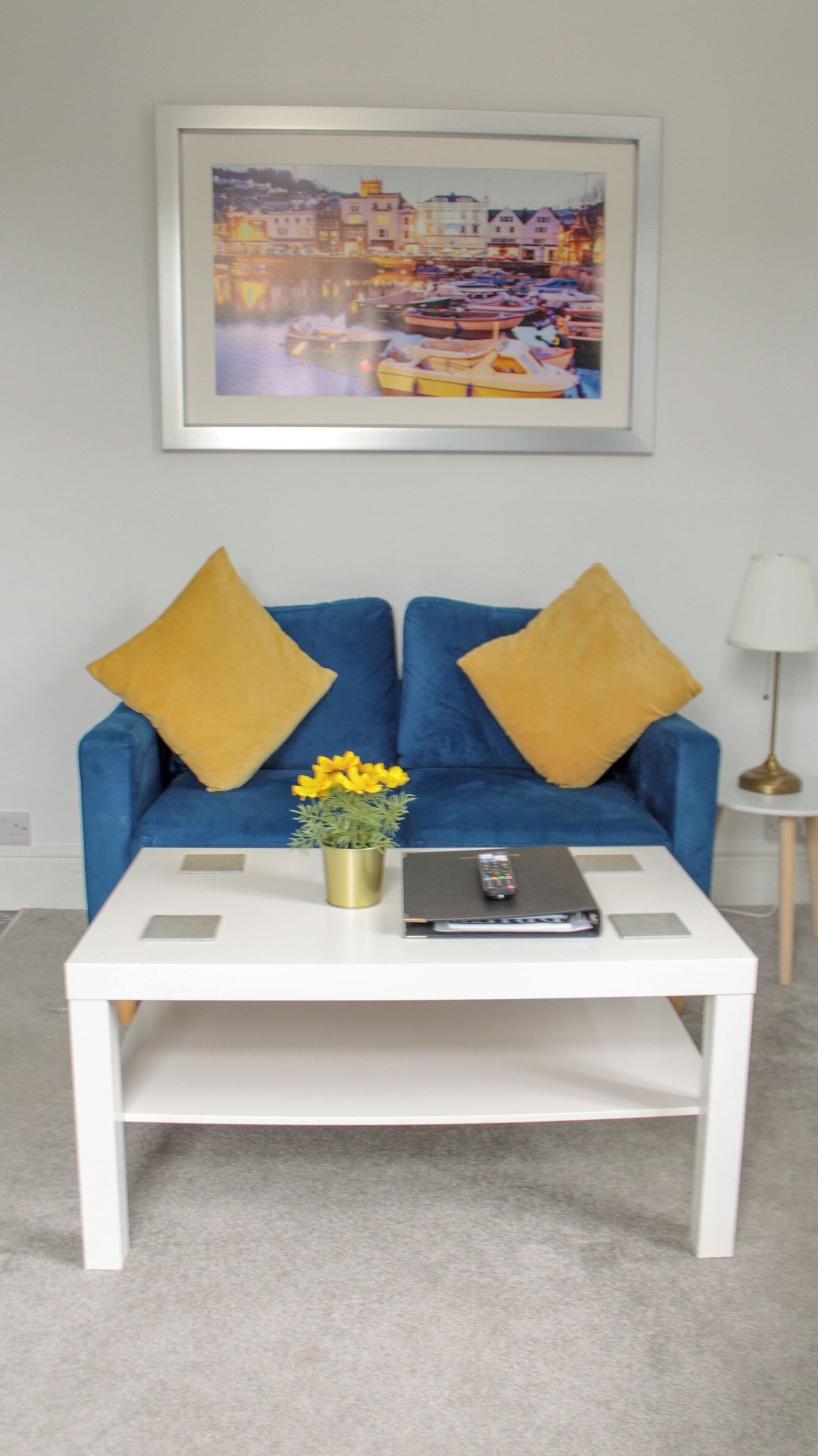 Uma sala de estar com um sofá azul, duas almofadas amarelas, uma mesa de centro branca, um quadro na parede e uma pequena luminária em uma mesinha lateral.