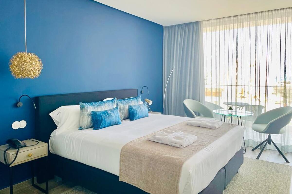 Quarto de hotel com uma cama grande, uma mesa de cabeceira com telefone, uma cadeira, uma pequena mesa e vista para o exterior.
