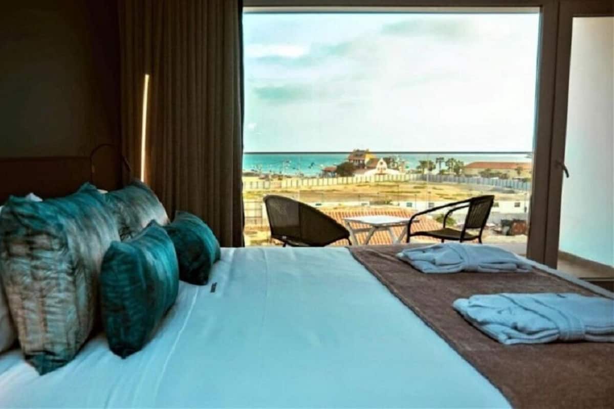 Quarto de hotel com cama, almofadas, uma cadeira e vista para a praia e edifícios.
