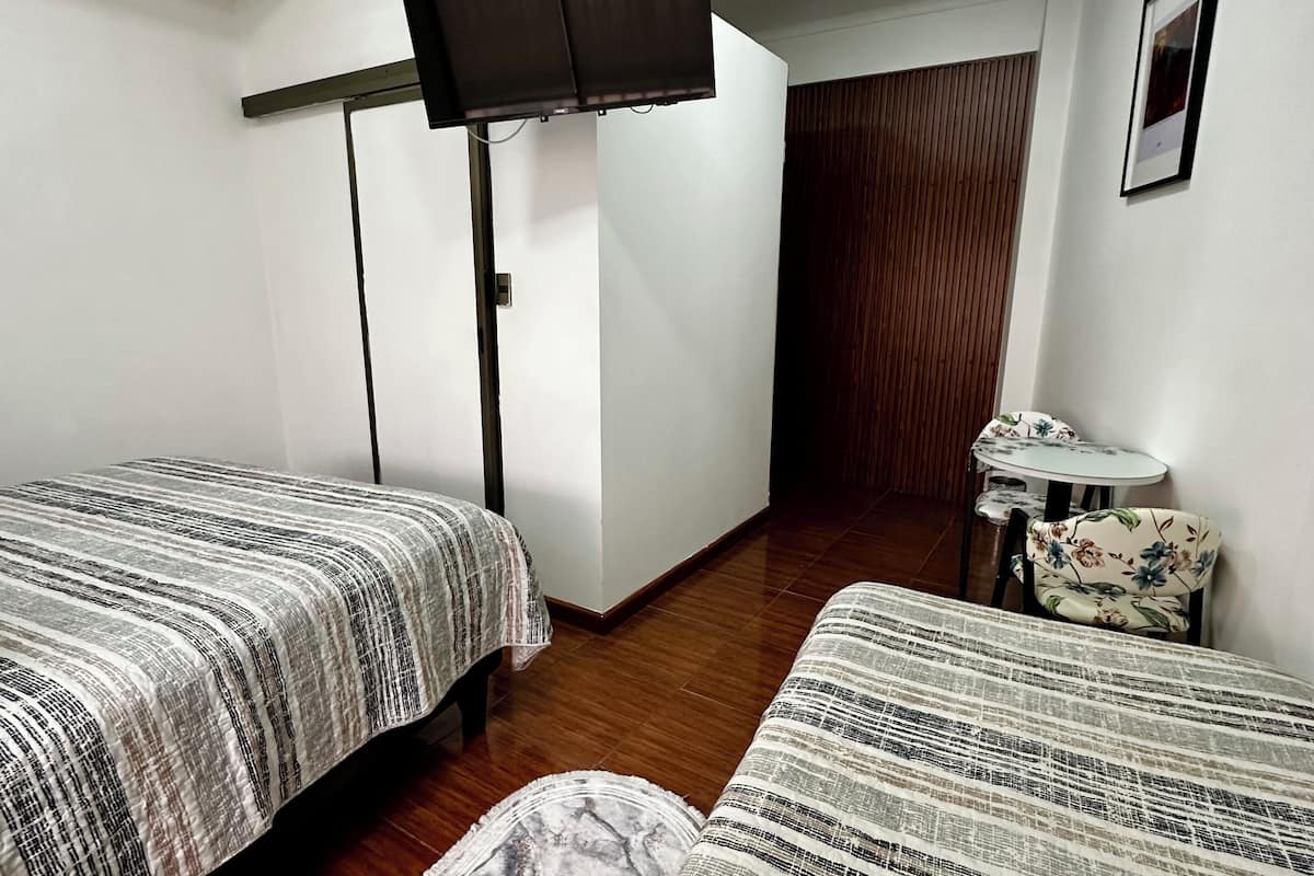 Quarto com duas camas, uma mesa pequena, televisão fixada na parede e uma porta de madeira.