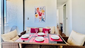 Uma sala de jantar com móveis de vime, uma mesa posta para quatro pessoas e um quadro floral na parede.
