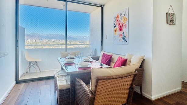 Uma área de refeições com mesa posta para quatro pessoas, uma cadeira de vime, um sofá com almofadas e uma janela grande com vista para a cidade.