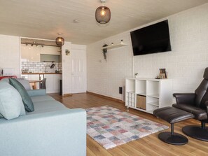 Uma sala de estar moderna com sofá, poltrona de couro, televisão e cozinha ao fundo.
