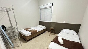 Quarto com duas camas, uma mesinha, janela com persianas e um suporte de metal.