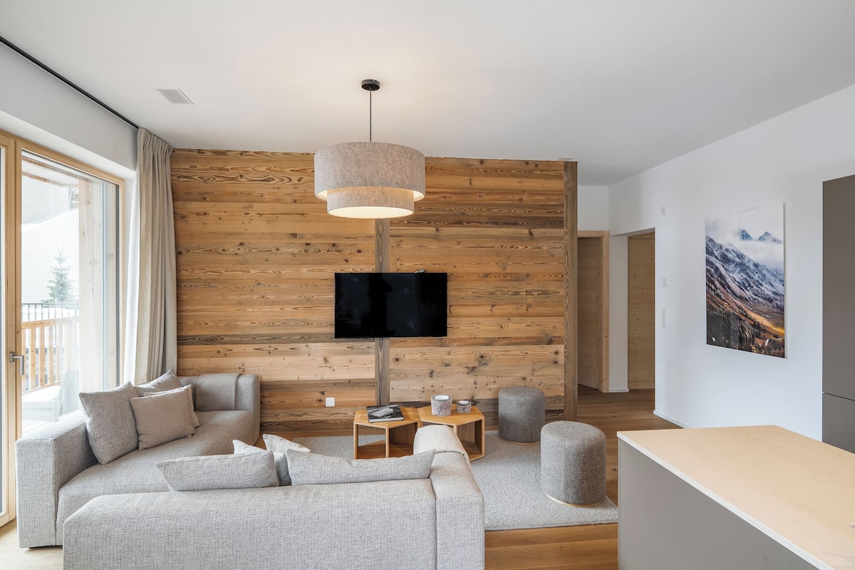 Uma sala de estar moderna com um sofá seccional, uma TV fixada em uma parede de madeira, uma luminária pendente e uma mesinha com duas velas.
