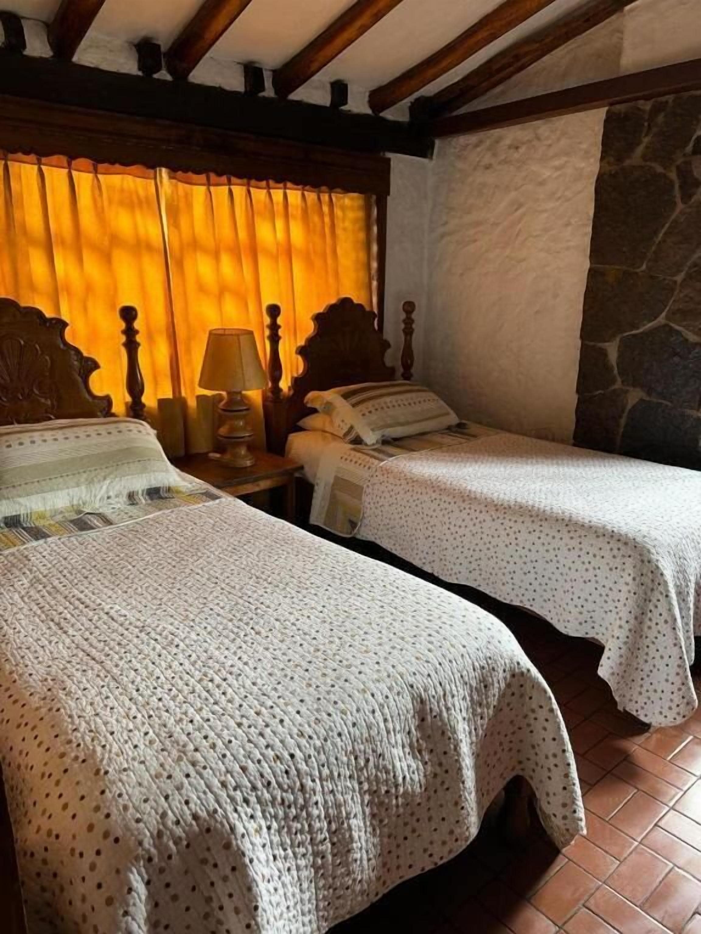 Quarto com duas camas, uma luminária e parede de pedra.