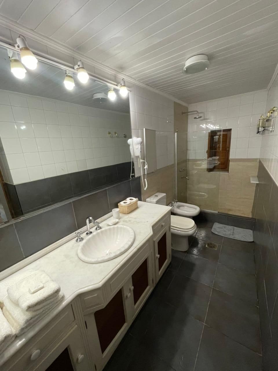 Casa de banho com toucador branco, espelho, chuveiro e sanita.