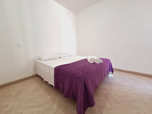 Uma cama de solteiro com colchão branco, cobertor roxo, travesseiro branco e duas toalhas enroladas sobre a cama.