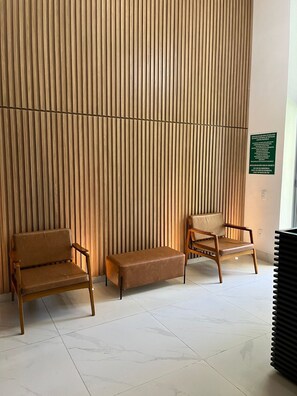 Uma área de recepção com um painel de madeira ripas, duas poltronas de couro, um banco e uma placa na parede.