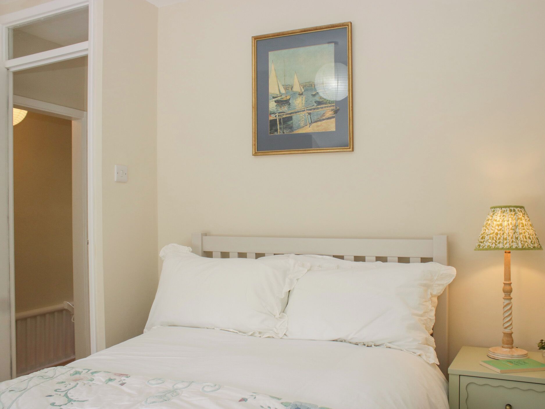 Quarto com cama, mesa de cabeceira com abajur, quadro na parede e porta para outro cômodo.