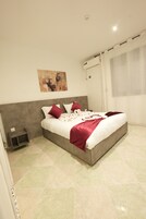 Quarto com cama, almofadas, cabeceira, mesa de cabeceira pequena, ar condicionado fixado na parede e janela com cortinas.