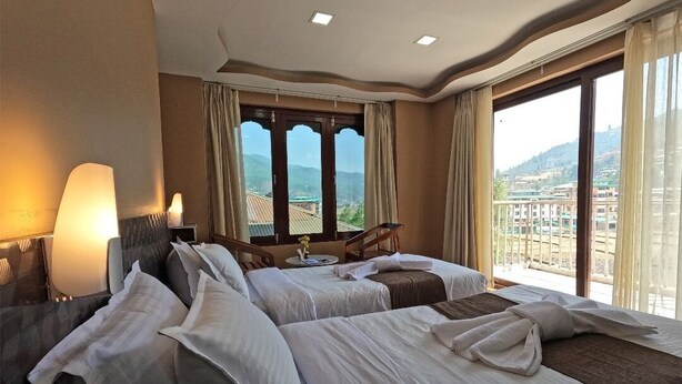 Quarto de hotel com duas camas, janela com vista para a cidade, luminária e pequena mesa com cadeira.