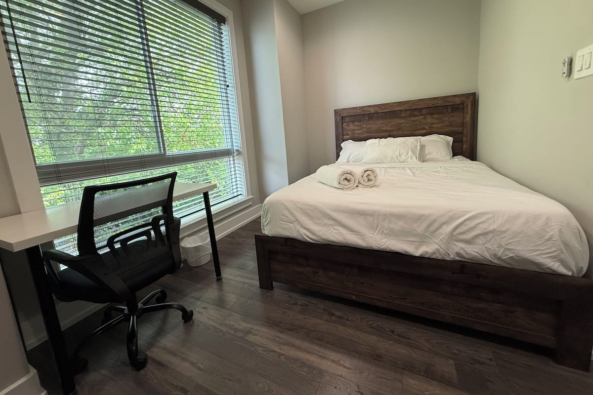 Um quarto com uma cama de madeira, uma escrivaninha com cadeira e uma janela com persianas.