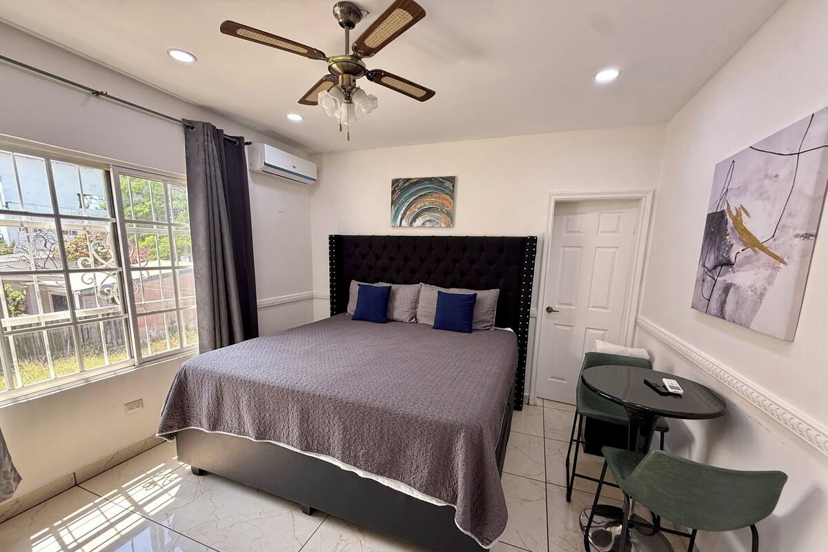 Um quarto com uma cama, ventilador de teto, janela com cortinas, porta, pequena mesa com cadeiras e obras de arte nas paredes.