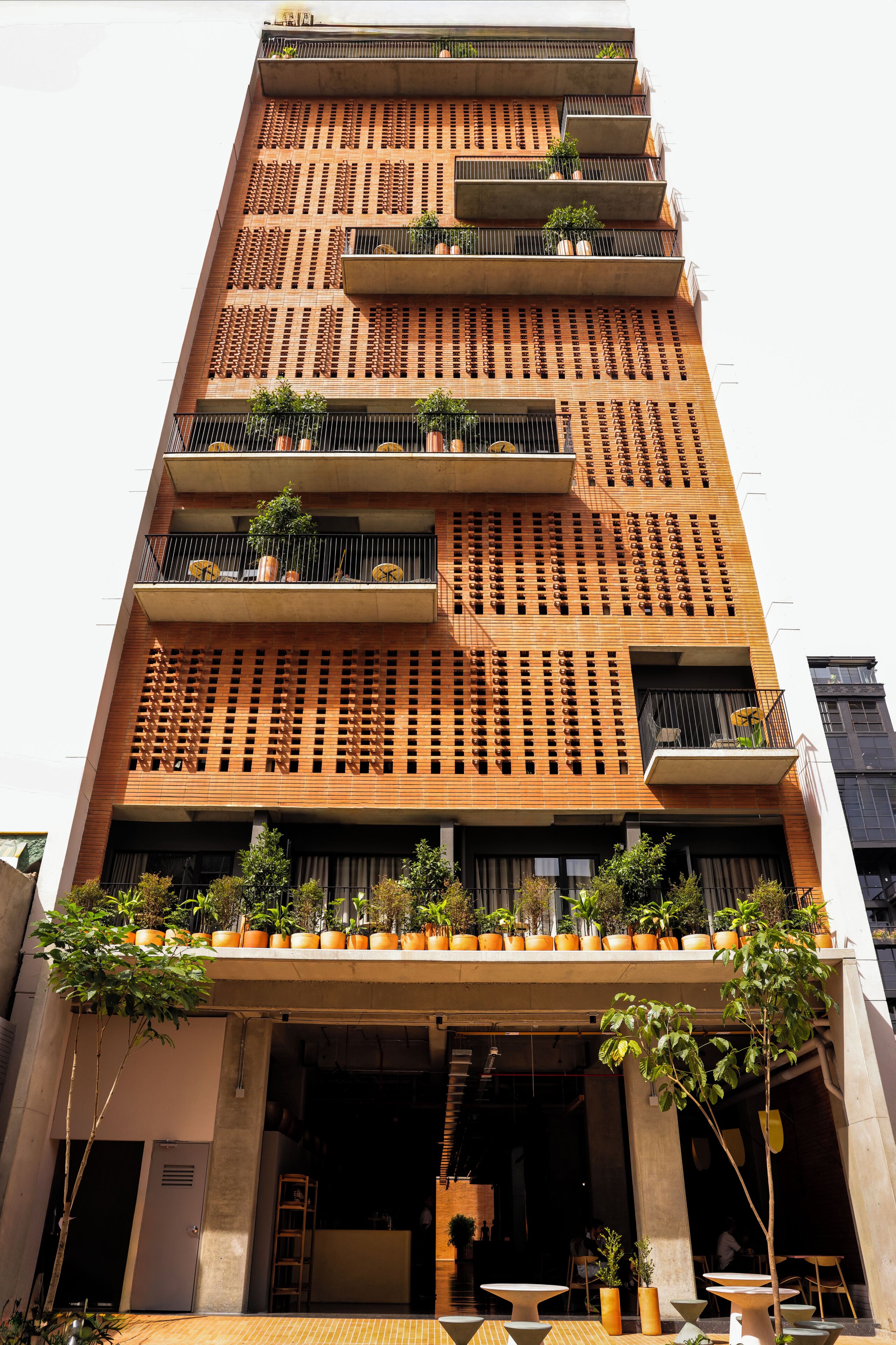 Edifício de vários andares com varandas, plantas em vasos e entrada no nível da rua com assentos.