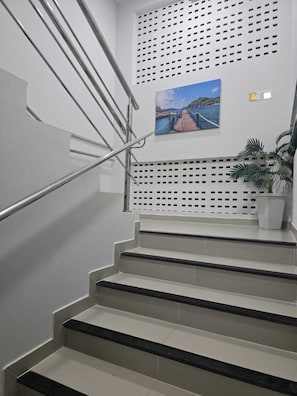 Uma escadaria com corrimão de metal, paredes brancas e uma imagem de uma paisagem costeira.