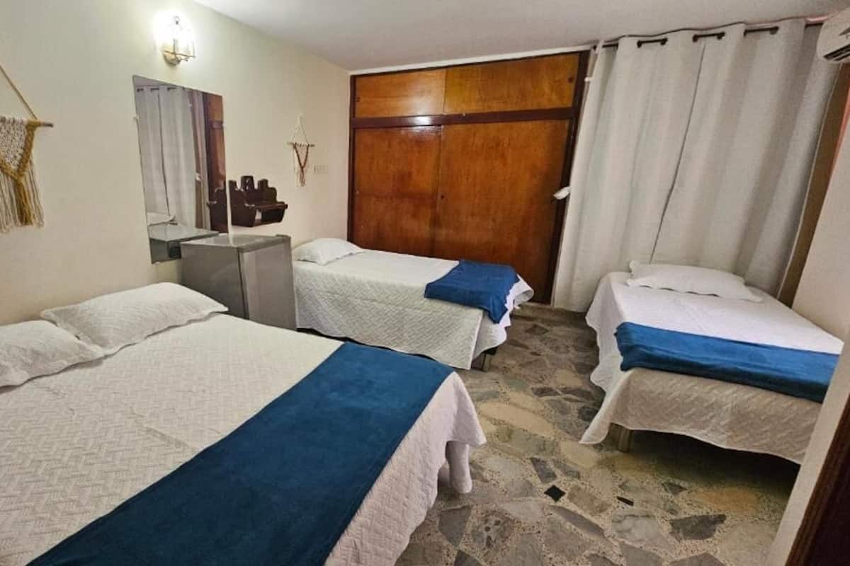 Quarto com três camas, um guarda-roupa de madeira, um espelho e um ornamento de macramé na parede.