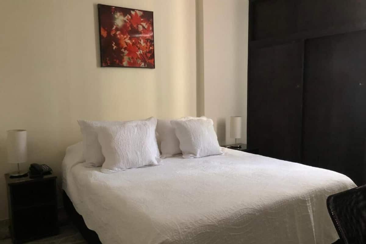 Uma cama bem arrumada com lençóis brancos, uma mesa de cabeceira com abajur e um guarda-roupa de cor escura.