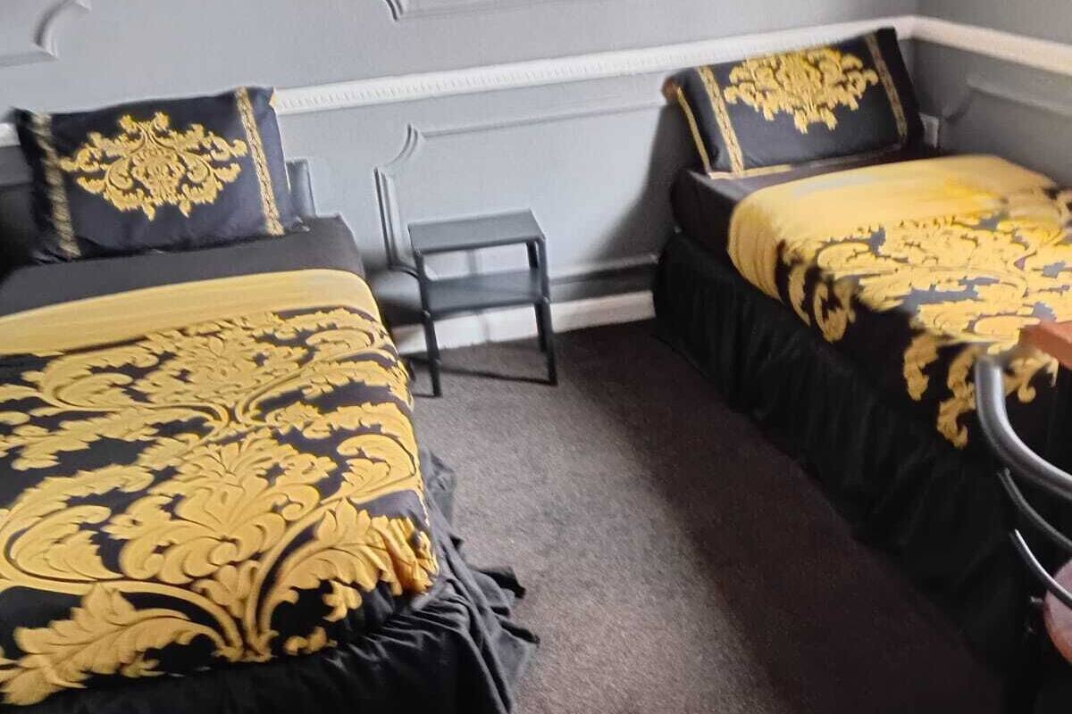 Quarto com duas camas, cada uma com colcha preta e dourada, uma cadeira preta pequena e uma parede cinza com tomada branca.