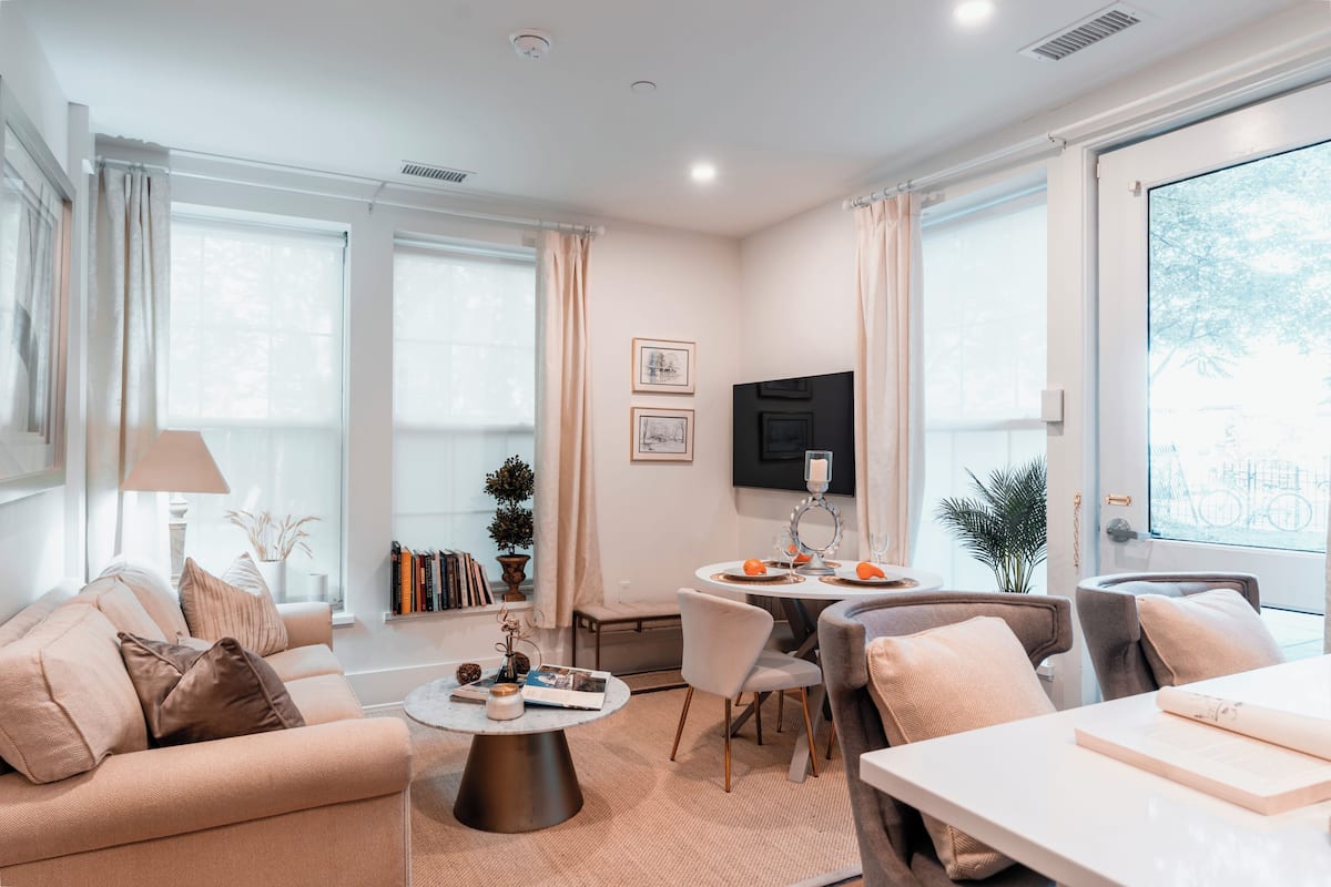 Uma sala de estar moderna com sofá, mesa de centro e área de refeições. Há janelas amplas com cortinas, uma televisão de tela plana e uma estante de livros.