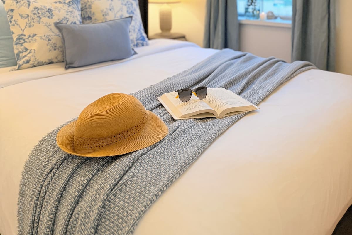 Uma cama bem arrumada com chapéu, óculos de sol e um livro aberto, um quadro na parede e uma janela com cortinas.