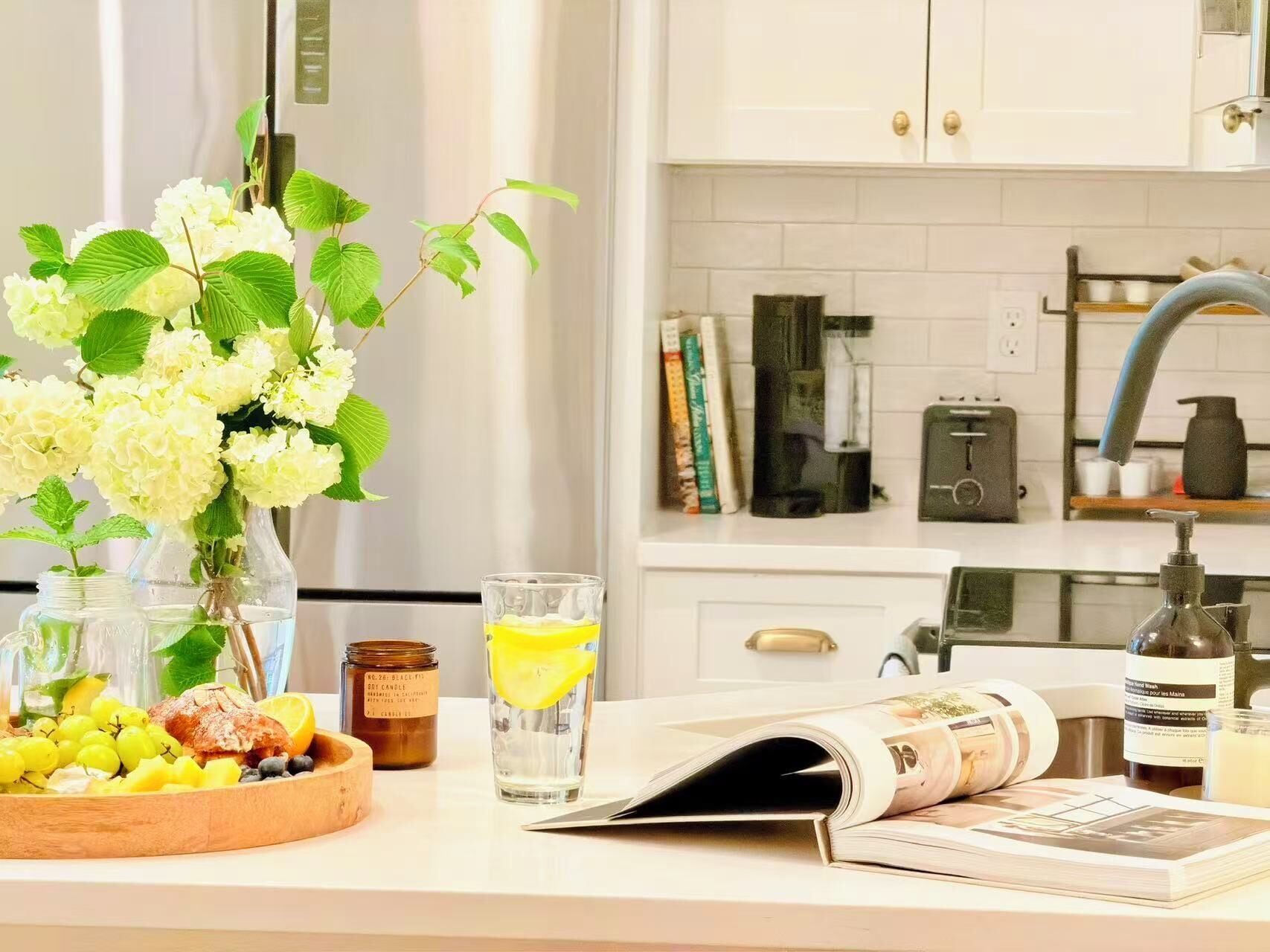 Uma bancada de cozinha com uma fruteira, um copo de água, uma vela e uma revista.