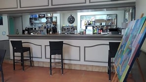 Uma área de bar com dois banquetas pretas, um relógio, prateleiras com garrafas e um quadro na parede.