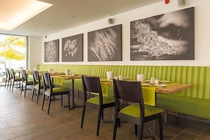 Um restaurante com cabines listradas de verde, mesas e cadeiras de madeira. Quatro grandes pinturas em preto e branco decoram a parede.