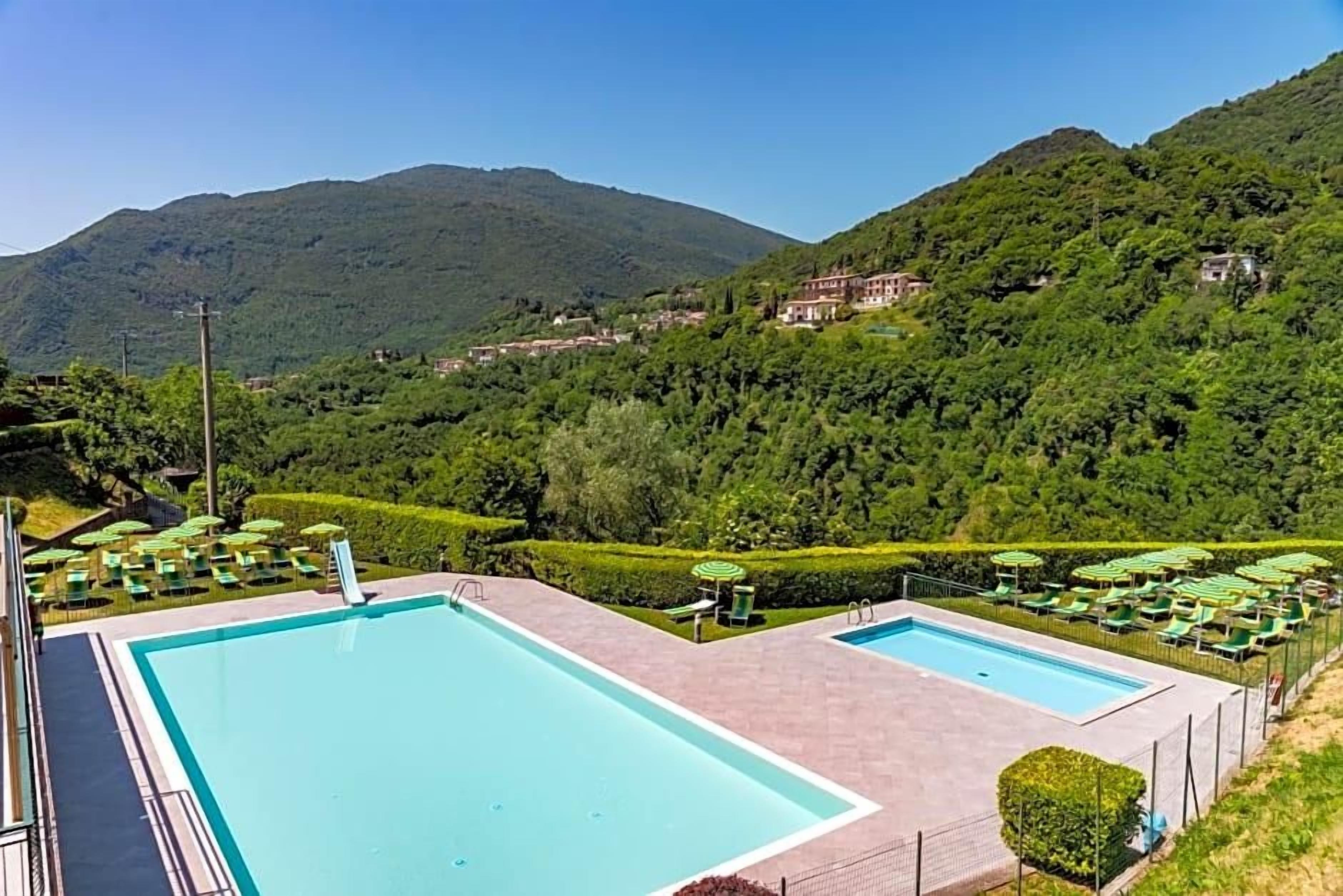 Uma área de piscina com duas piscinas retangulares, rodeada por guarda-sóis verdes e espreguiçadeiras, com vista para colinas verdes exuberantes e um céu limpo.