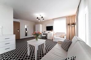 Quarto de hotel com carpete estampado, sofá branco, uma pequena mesa com um vaso de flores, escrivaninha com cadeira, televisão e guarda-roupa.