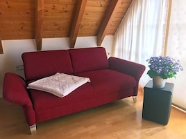 Um sofá vermelho com uma almofada branca numa sala com vigas de madeira, uma planta em vaso e uma janela com cortinas.