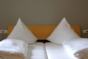 Cama com lençóis brancos e duas almofadas, cabeceira de madeira e uma tomada na parede.