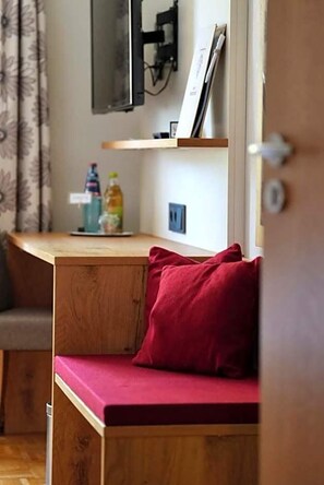 Quarto com escrivaninha de madeira, almofada vermelha, TV na parede e prateleira com garrafas.