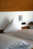 Quarto de hotel com teto de madeira, cama com roupa de cama branca, mesa de cabeceira com abajur e termostato fixado na parede.