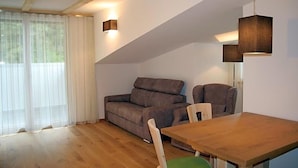 Quarto com piso de madeira, sofá, mesa de jantar e janela com cortinas leves.