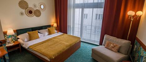 Quarto de hotel com cama, cadeira, mesa de cabeceira, luminária e janela com cortinas.