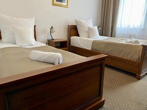 Quarto de hotel com duas camas de madeira, cada uma com roupa de cama e almofadas brancas. Há uma pequena mesa de cabeceira com uma luminária, e o chão do quarto é coberto por carpete.