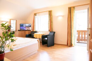 Quarto de hotel com cama, escrivaninha com televisão, cadeira, mesinha e varanda com cortinas.