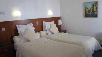 Quarto de hotel com cabeceira de madeira, duas camas com roupa de cama branca, mesa de cabeceira com abajur e um quadro na parede.
