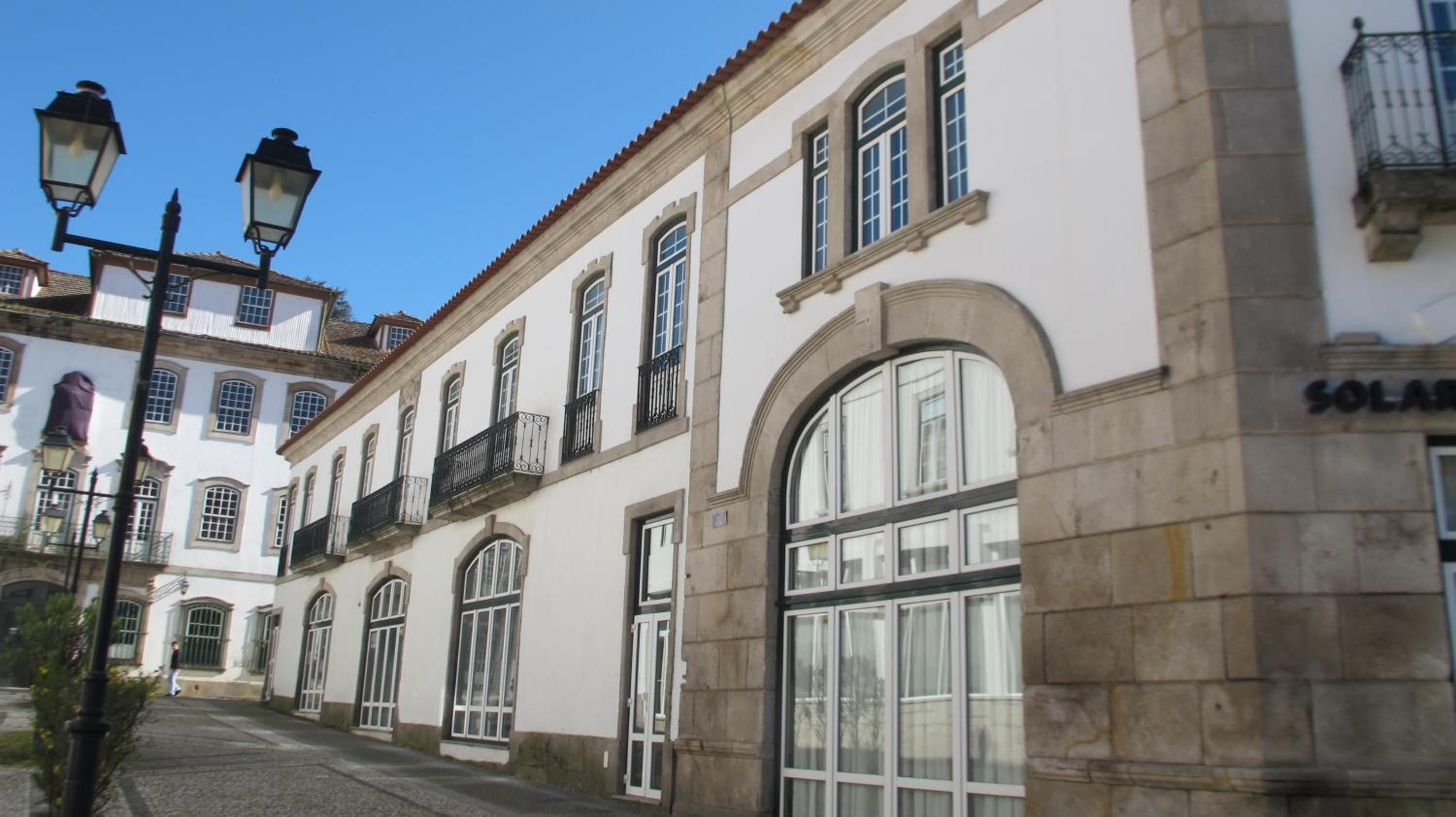 Uma rua com edifícios com janelas em arco e varandas, um caminho de paralelepípedos e luminárias.