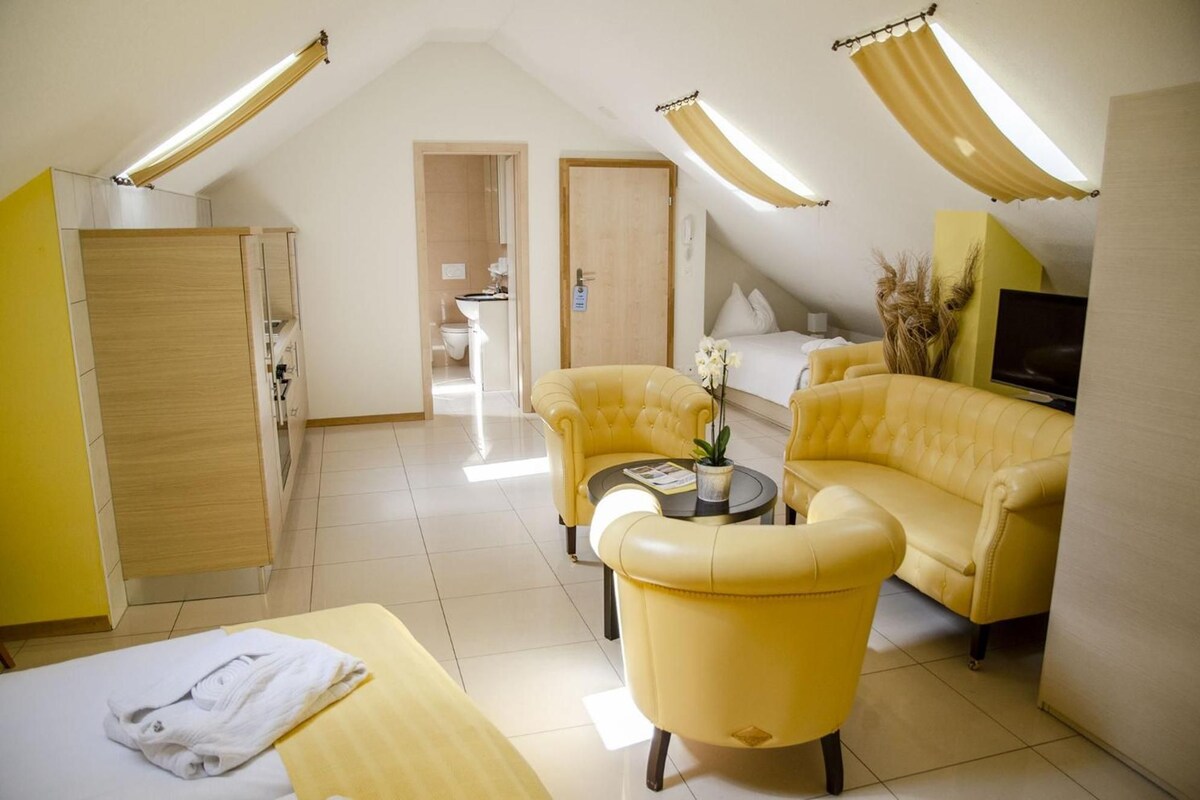Quarto com cama, sofá amarelo, uma mesinha e televisão. Uma porta dá acesso a uma casa de banho.