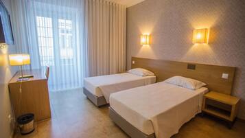 Quarto de hotel com duas camas, uma escrivaninha, uma cadeira, um abajur e uma janela com cortinas.