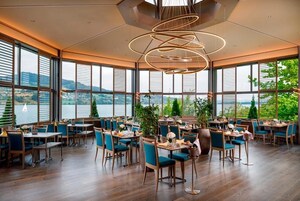 Um restaurante com várias mesas e cadeiras, amplas janelas com vista para a água e um lustre moderno.