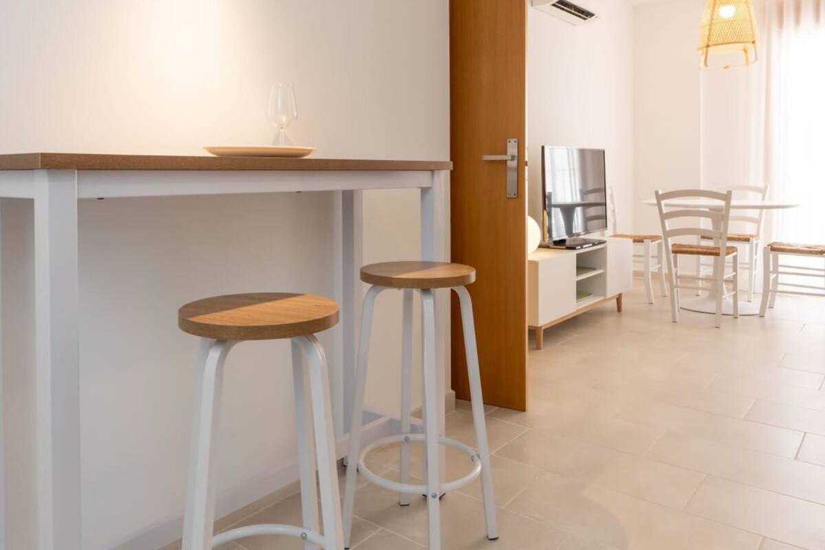 Cozinha moderna com bancada de madeira, dois bancos brancos, uma mesa de jantar com cadeiras e uma TV.