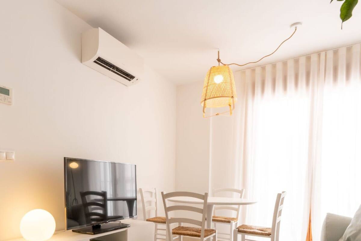 Uma sala de estar moderna com uma televisão de tela plana, um rack branco para equipamentos eletrônicos, uma mesa de jantar com cadeiras e um lustre.