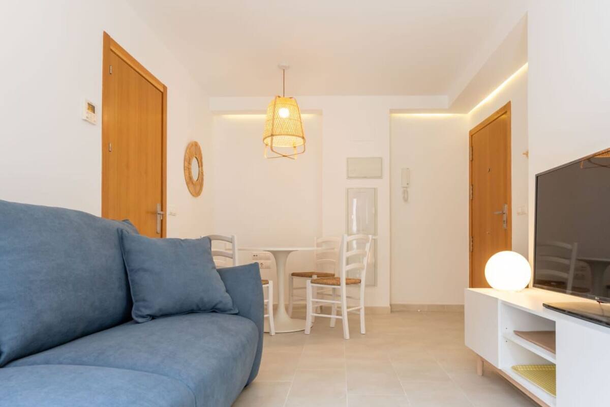 Uma sala de estar moderna com um sofá, uma mesa de jantar com cadeiras, uma TV sobre um móvel e um lustre.