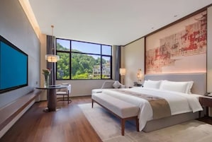 Quarto de hotel moderno com cama grande, escrivaninha, cadeira e ampla janela com vista para áreas verdes e edifícios.