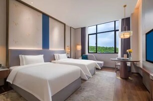 Quarto de hotel com duas camas, uma escrivaninha, uma cadeira, uma mesa redonda com um vaso e uma grande janela com vista para o verde.